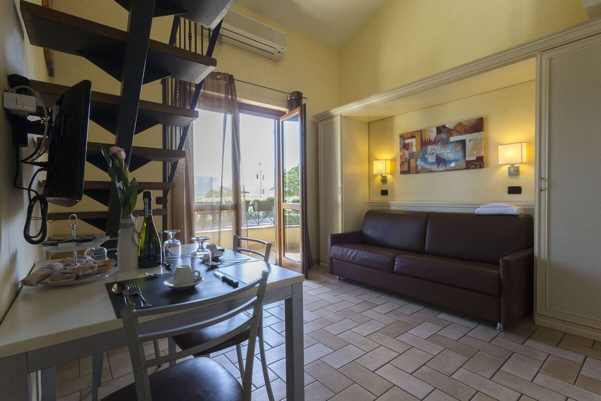 Uma área de refeições com uma mesa posta para dois, um sofá de couro marrom e uma varanda com vista.