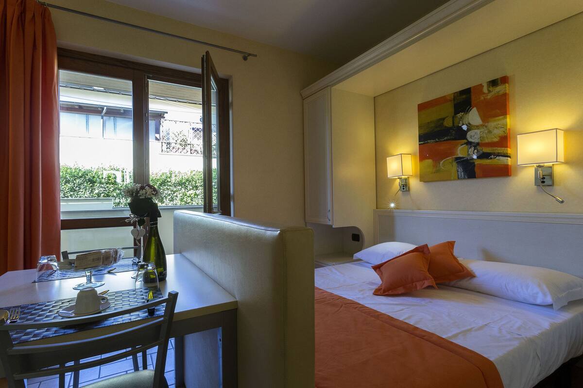 Quarto de hotel com uma cama, uma mesa com cadeira, uma janela com cortinas e um quadro na parede.