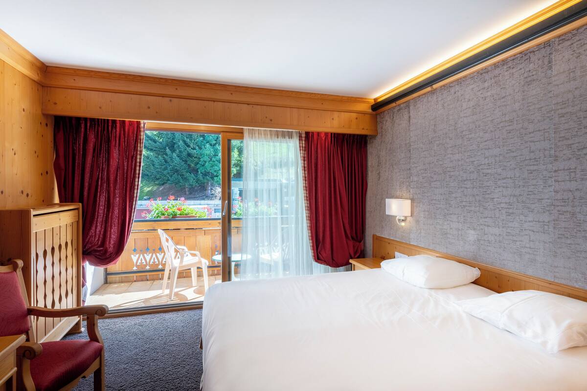Quarto de hotel com cama, cabeceira de madeira, escrivaninha de madeira, uma cadeira e vista através de uma porta de vidro para área externa com plantas.