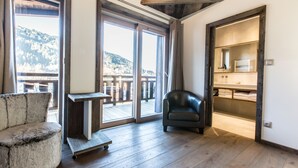 Quarto com piso de madeira, uma poltrona de couro, uma mesa pequena e vista para uma varanda com guarda-corpo.