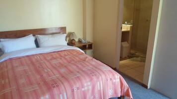 Quarto de hotel com uma cama, uma mesa de cabeceira, uma luminária e um banheiro visível através de uma porta aberta.