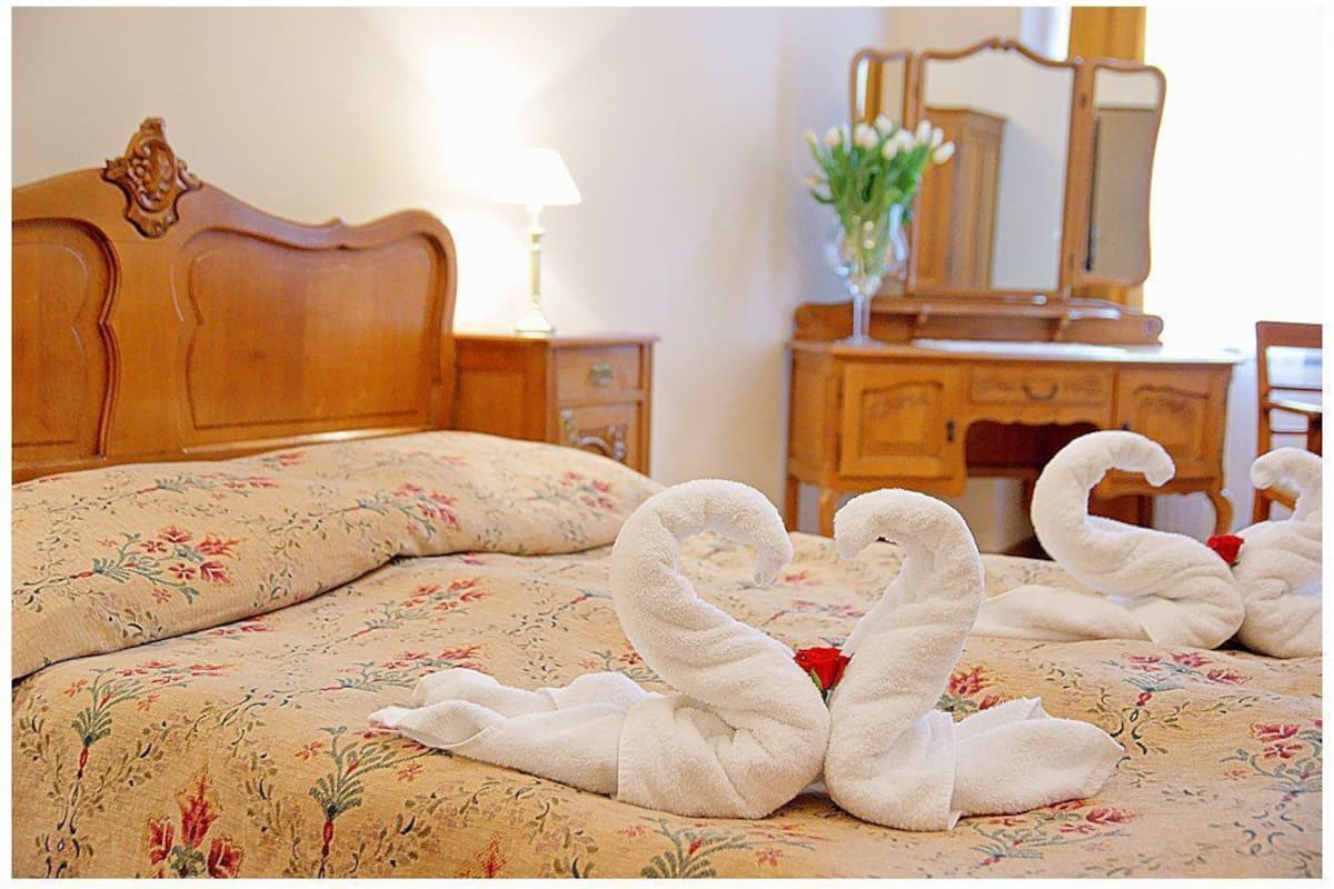 Quarto de hotel com colcha estampada em flores, cabeceira de madeira, mesa de cabeceira com abajur, cômoda e espelho.