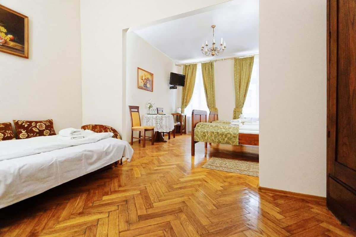 Quarto com piso de madeira em padrão herringbone, uma cama com colcha branca, uma mesinha com uma cadeira e uma televisão na parede.