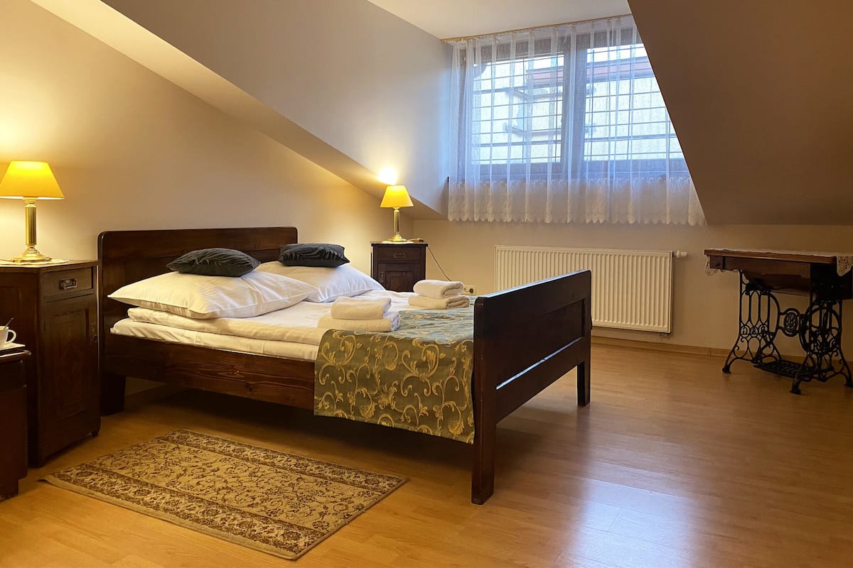 Quarto com cama, duas luminárias de cabeceira, radiador, mesa de madeira e janela com vista.