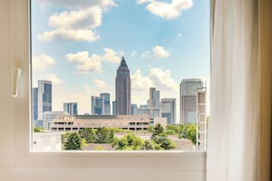 Vista de um horizonte urbano através de uma janela, destacando um arranha-céu, diversos edifícios altos e um estacionamento.