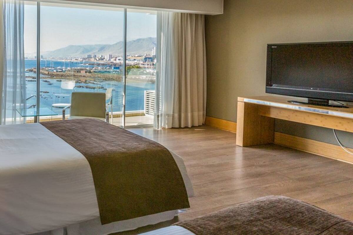 Quarto de hotel com cama, televisão, secretária e vista para uma cidade costeira.