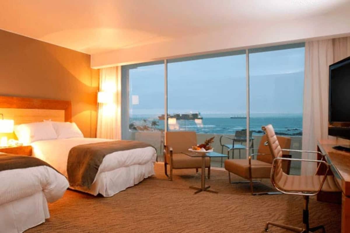 Quarto de hotel com duas camas, televisão, secretária e vista para o oceano.