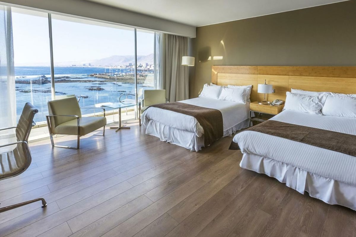 Quarto de hotel com duas camas, vista para o mar e varanda com uma cadeira.