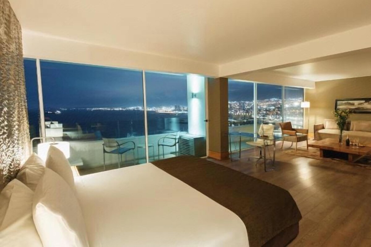Quarto de hotel moderno com uma cama grande, área de estar com sofá e cadeiras, e varanda com vista para a cidade à noite.