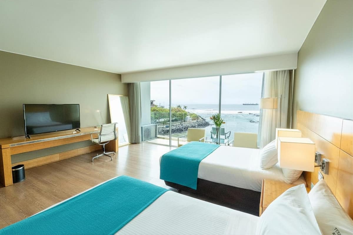 Quarto de hotel com duas camas, televisão, escrivaninha e vista para o oceano.
