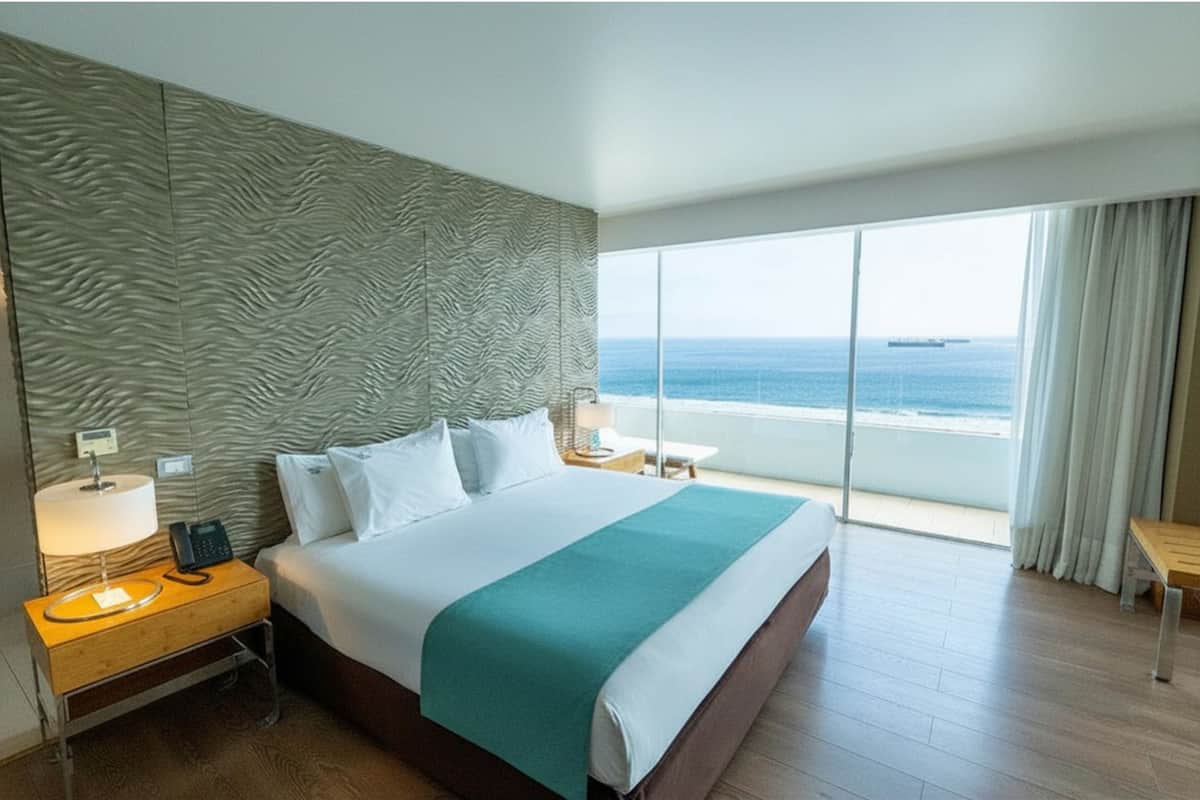 Quarto de hotel com uma cama grande, mesa de cabeceira com abajur e telefone, vista para o oceano através de janelas amplas e piso de madeira.