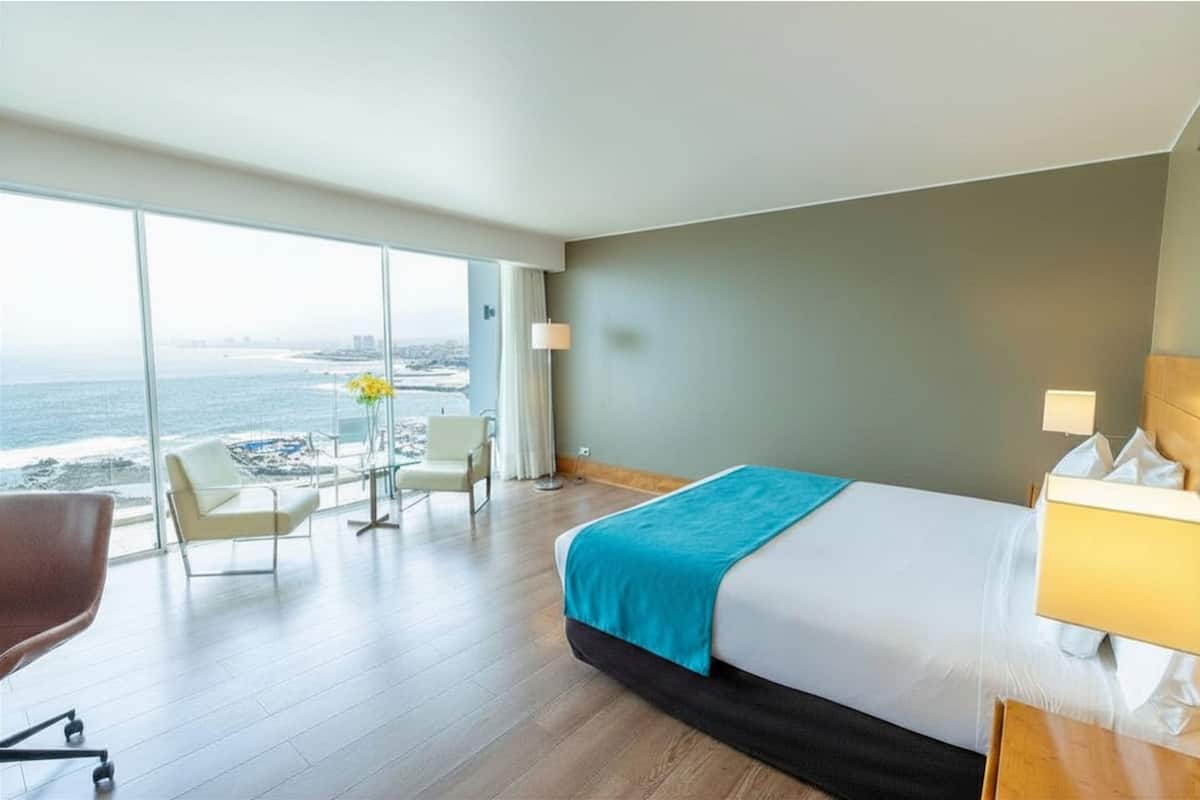Quarto de hotel com uma cama grande, uma escrivaninha, uma cadeira e vista para o oceano.