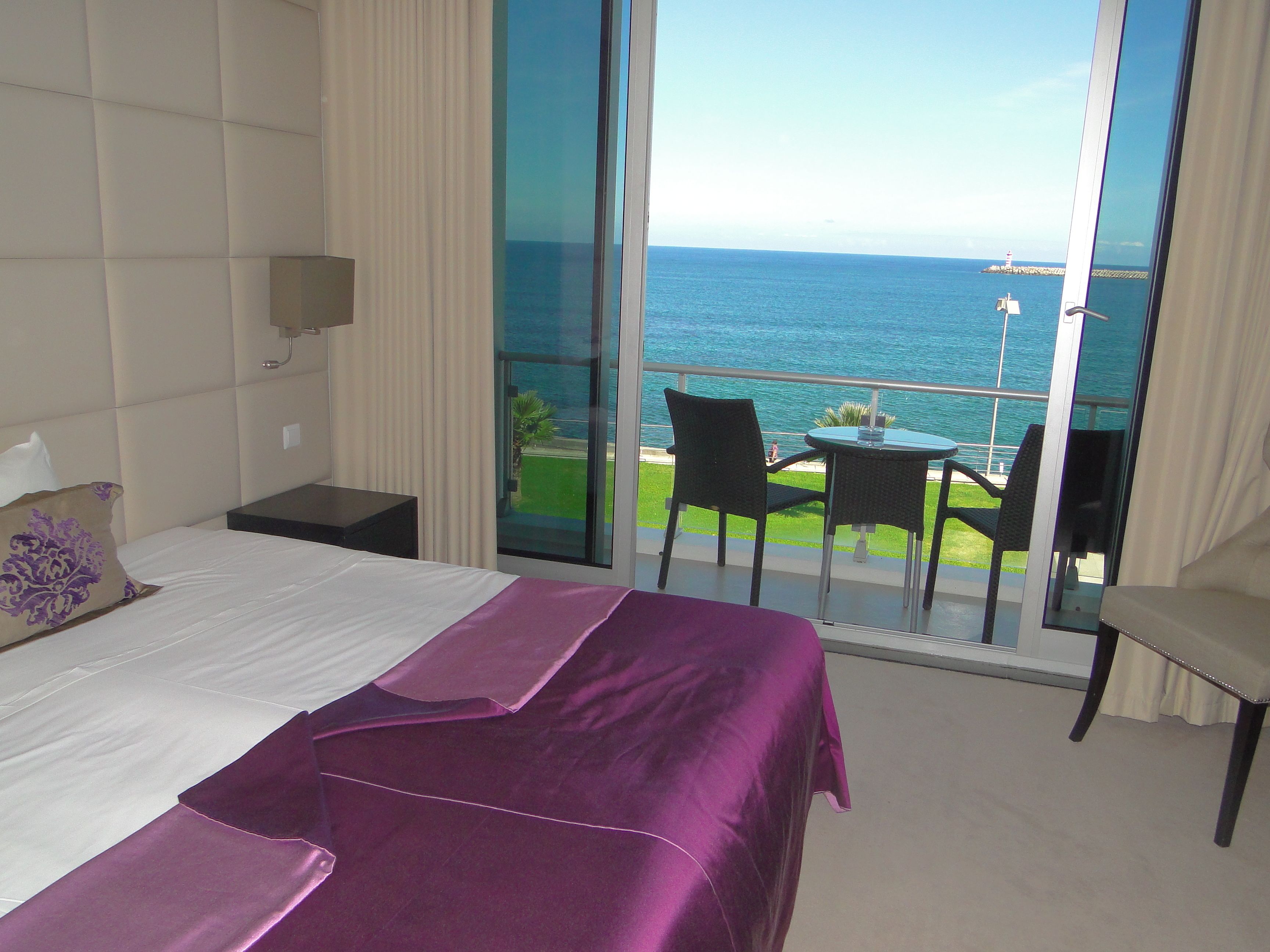 Quarto de hotel com cama, uma mesa pequena, duas cadeiras e vista para o oceano.