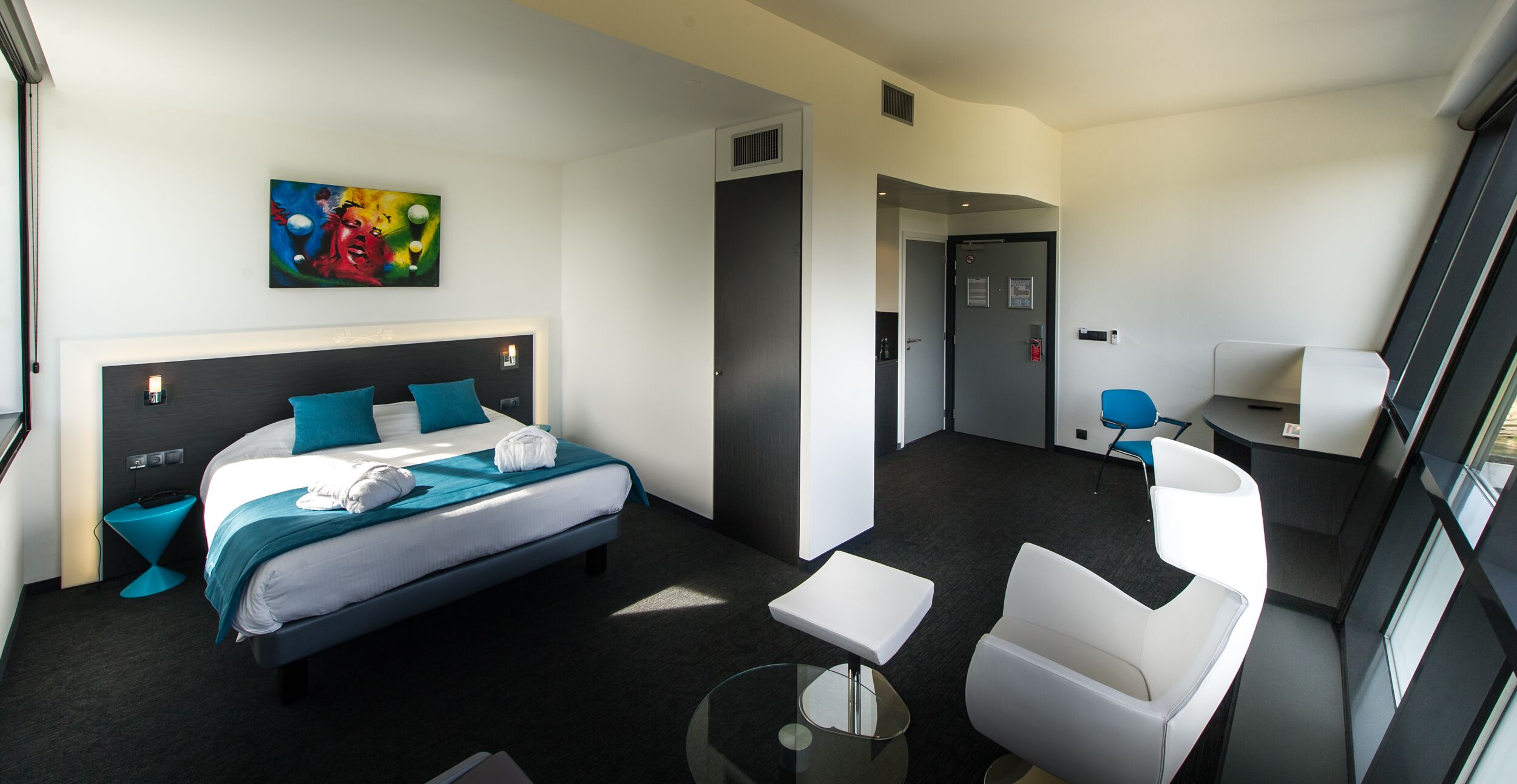 Quarto de hotel moderno com cama, escrivaninha, cadeira e um quadro na parede.