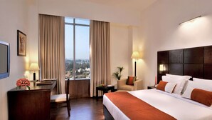 Quarto de hotel com uma cama grande, uma mesa de escritório, uma cadeira, mesa de cabeceira e vista para a cidade através de grandes janelas.