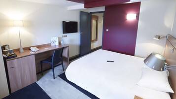 Quarto de hotel com cama, escrivaninha, cadeira, abajur e televisão.