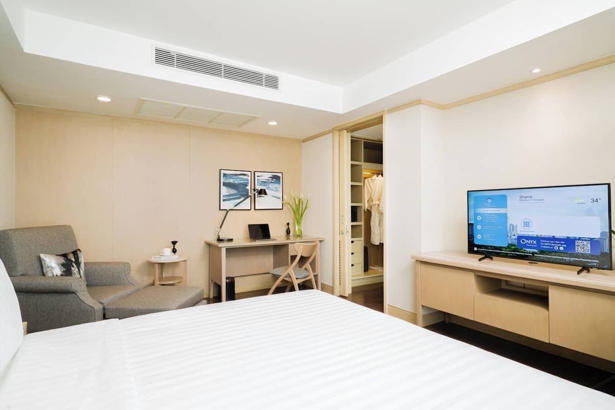 Quarto de hotel com cama, escrivaninha, cadeira, televisão e guarda-roupa.