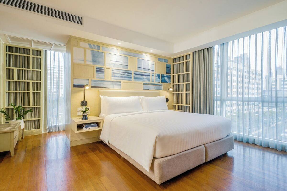 Quarto de hotel com uma cama grande, uma mesa de cabeceira, um abajur, uma planta e vista da cidade através de grandes janelas.