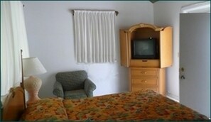 Quarto com cama, uma cadeira, televisão sobre um móvel de madeira e uma janela com cortinas.