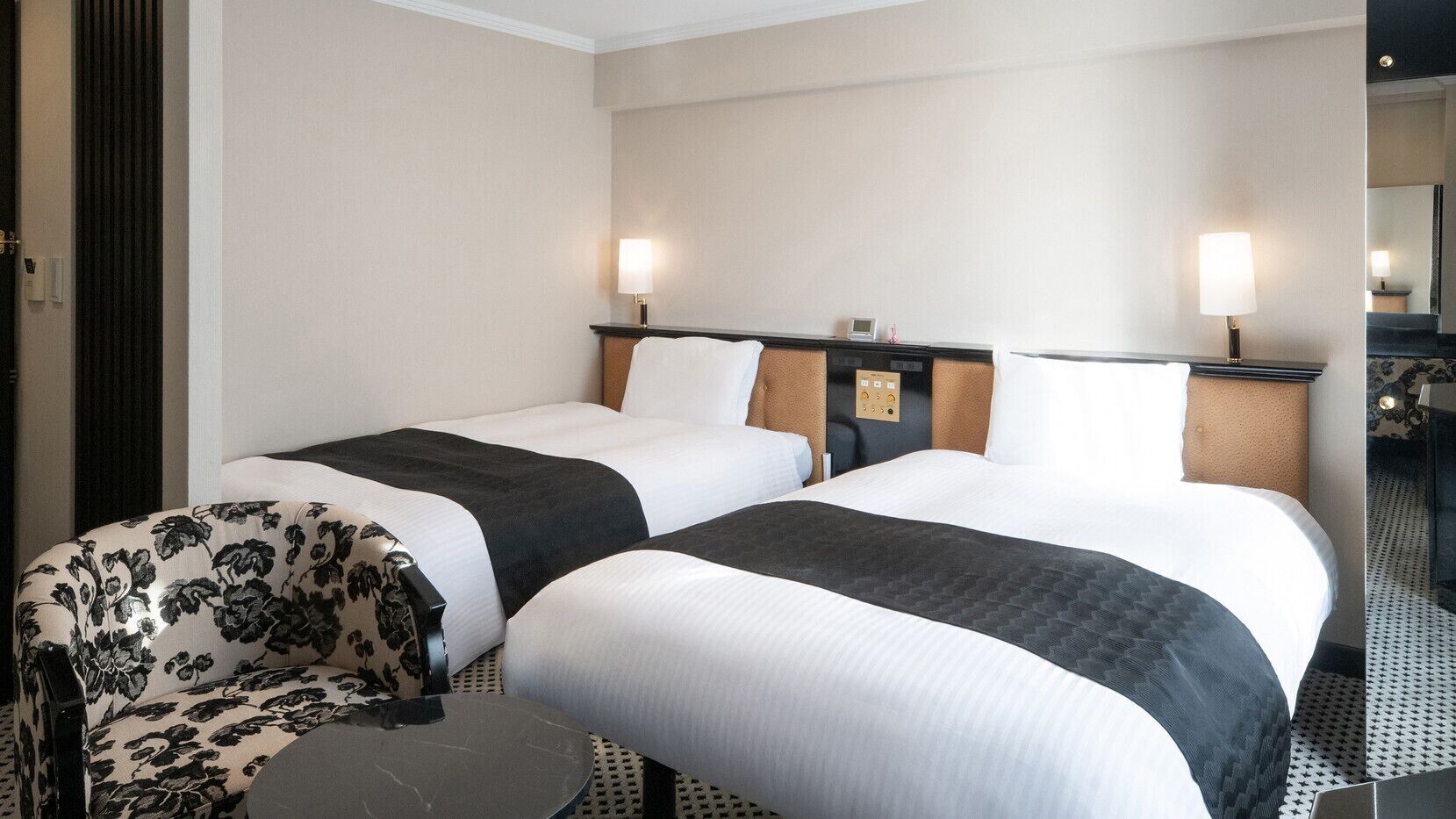 Quarto de hotel com duas camas, uma mesinha, uma cadeira e uma mesa de cabeceira com abajur.