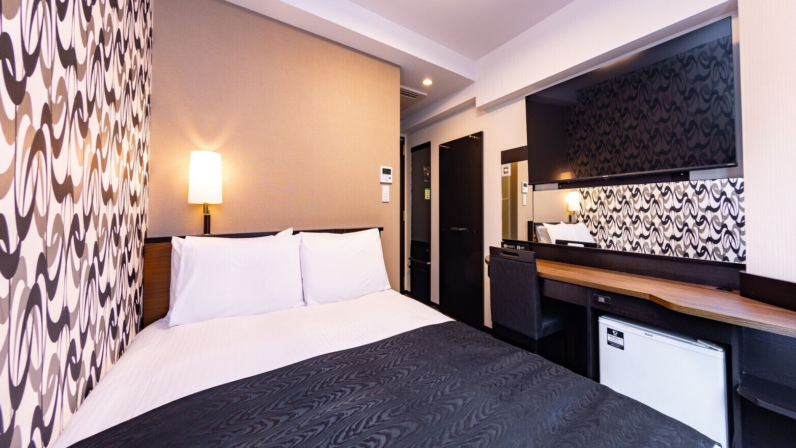 Quarto de hotel com cama, mesa de cabeceira, abajur e uma pequena cozinha equipada.