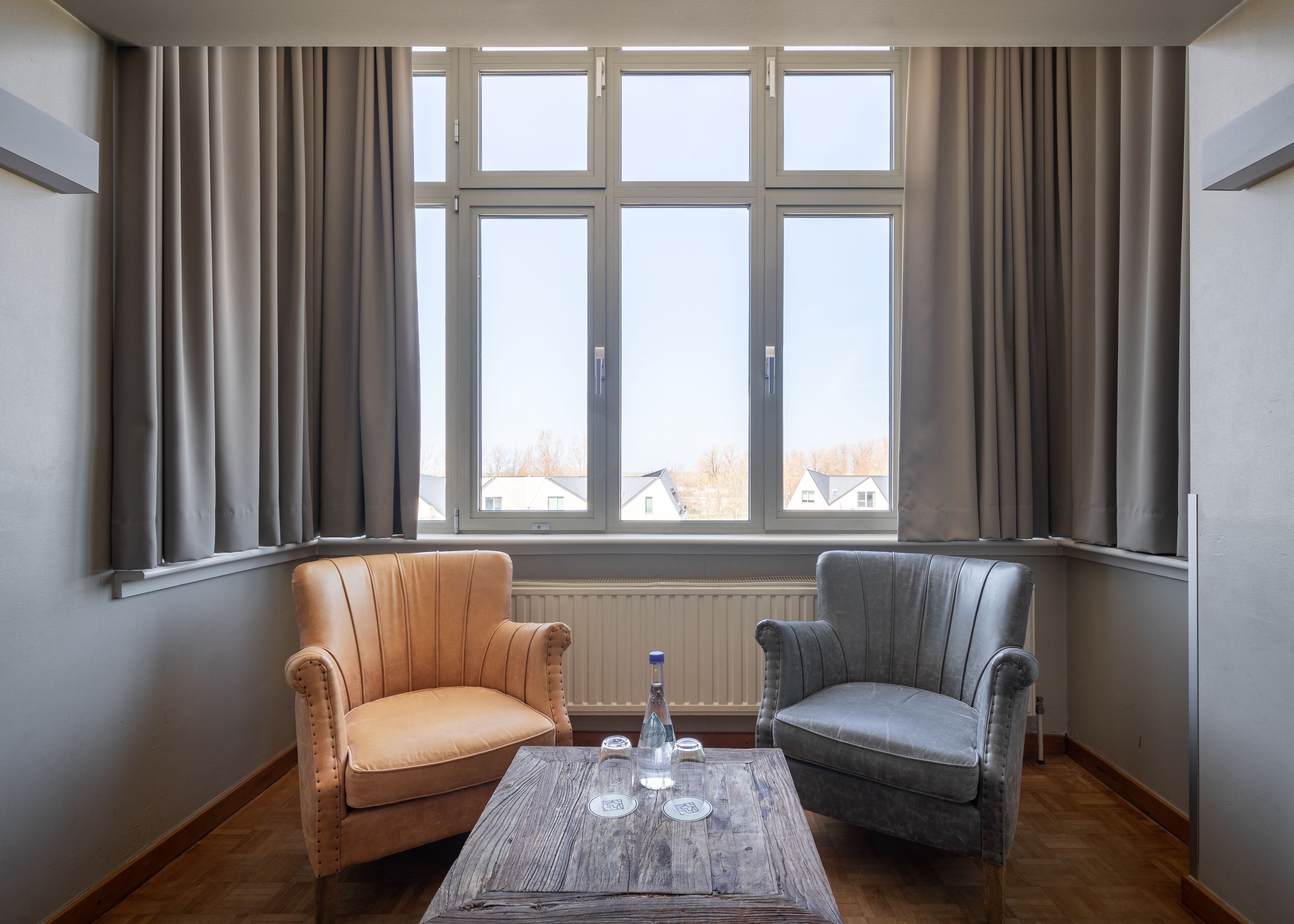 Quarto com duas poltronas de couro, uma mesa de centro de madeira com garrafas de água e uma janela grande com cortinas.