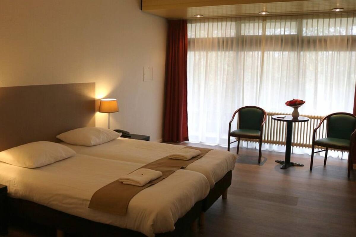 Quarto de hotel com uma cama grande, duas mesas de cabeceira com luminárias, uma pequena mesa redonda com duas cadeiras, e vista através de cortinas leves.