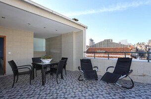 Uma área de estar ao ar livre com uma mesa, cadeiras, um vaso de planta e vista para edifícios ao fundo.