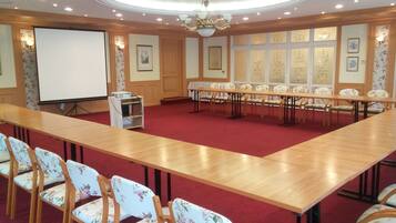 Uma sala de conferência com uma longa mesa de madeira, cadeiras estofadas com padrão floral, tela de projetor e quadros nas paredes.