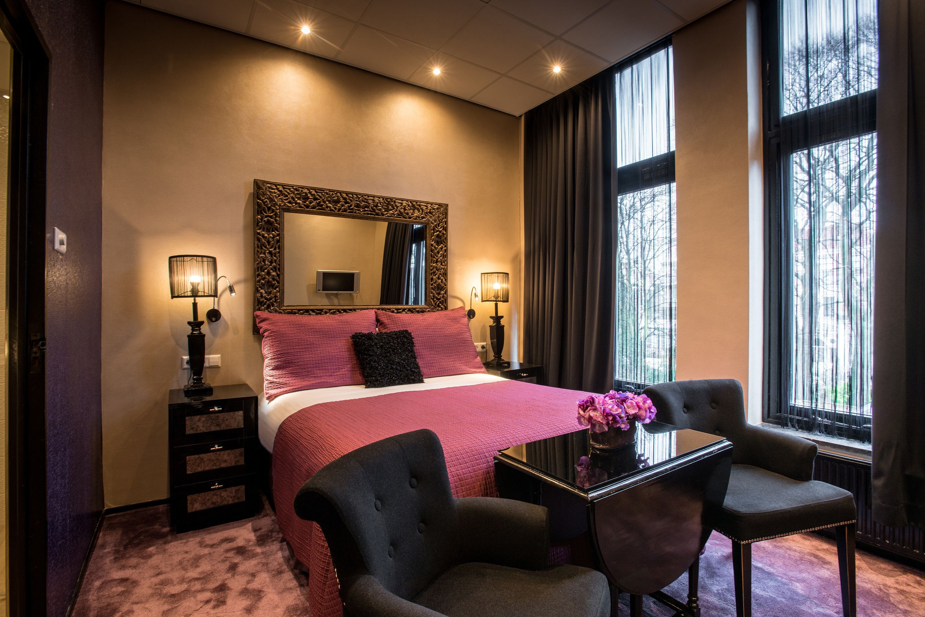 Quarto de hotel com uma cama grande, duas mesas de cabeceira com luminárias, uma escrivaninha com duas cadeiras e uma janela ampla com cortinas.