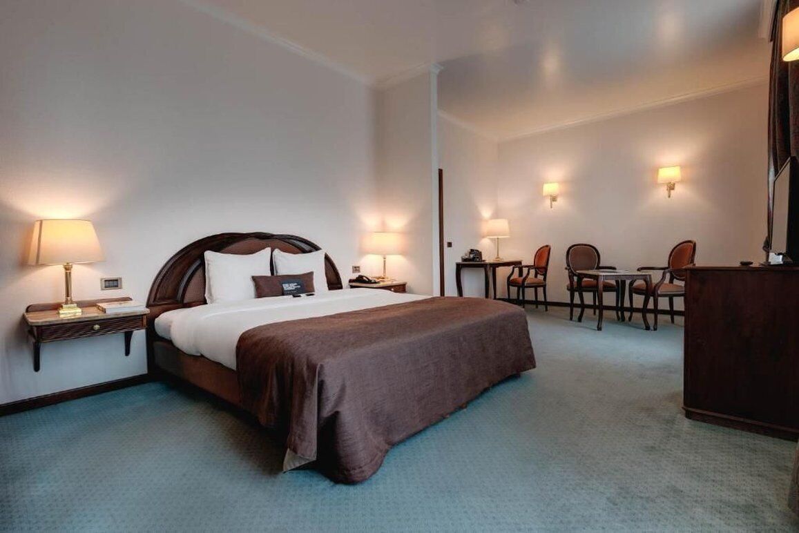 Quarto de hotel com cama, mesas de cabeceira, luminárias, escrivaninha com cadeiras e guarda-roupa.