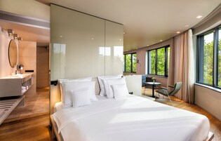 Quarto de hotel moderno com uma cama grande, uma escrivaninha e uma cadeira, banheiro com espelho e prateleiras, e grandes janelas com vista para árvores.