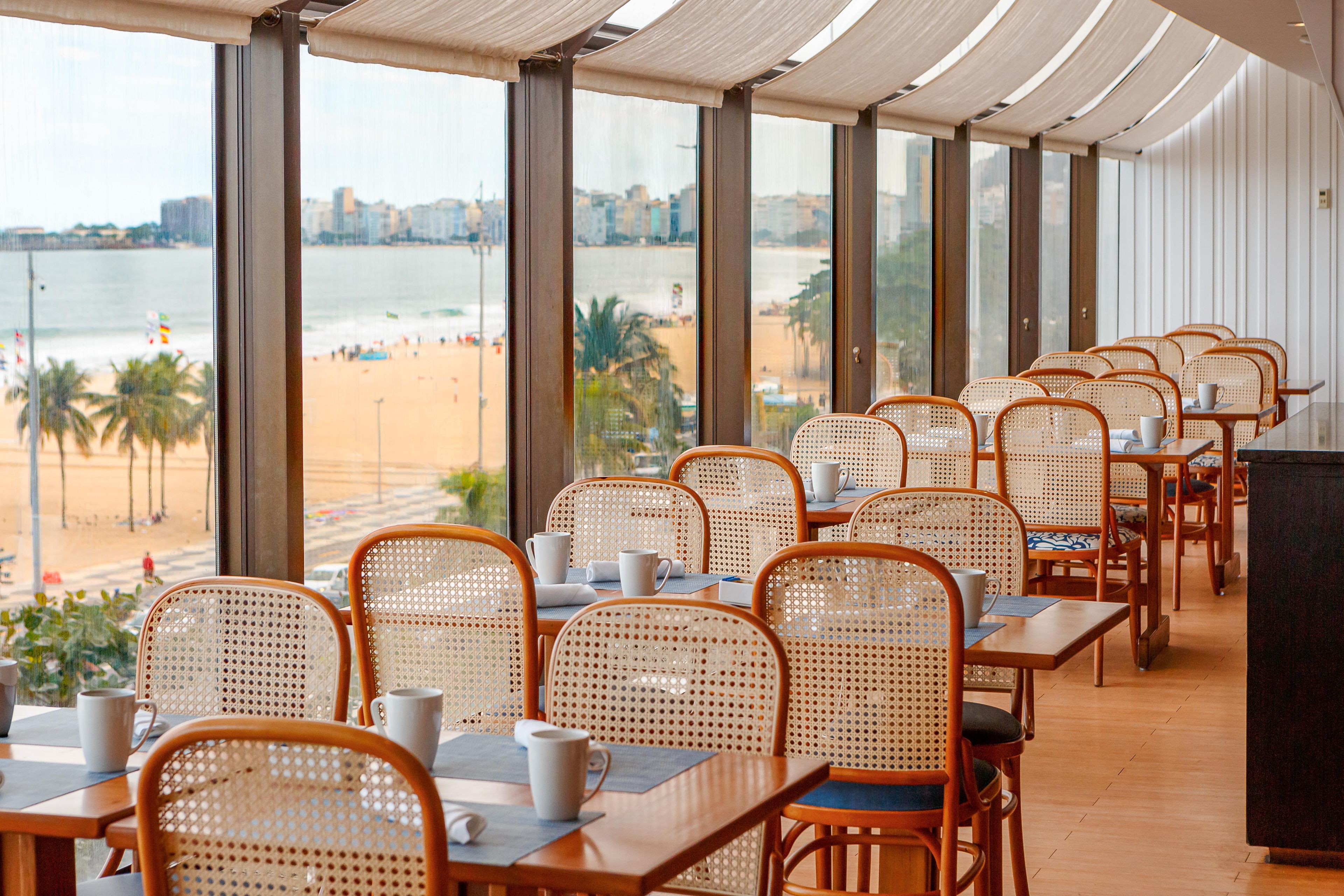 Restaurante à beira-mar com cadeiras e mesas de vime, e amplas janelas com vista para a praia.