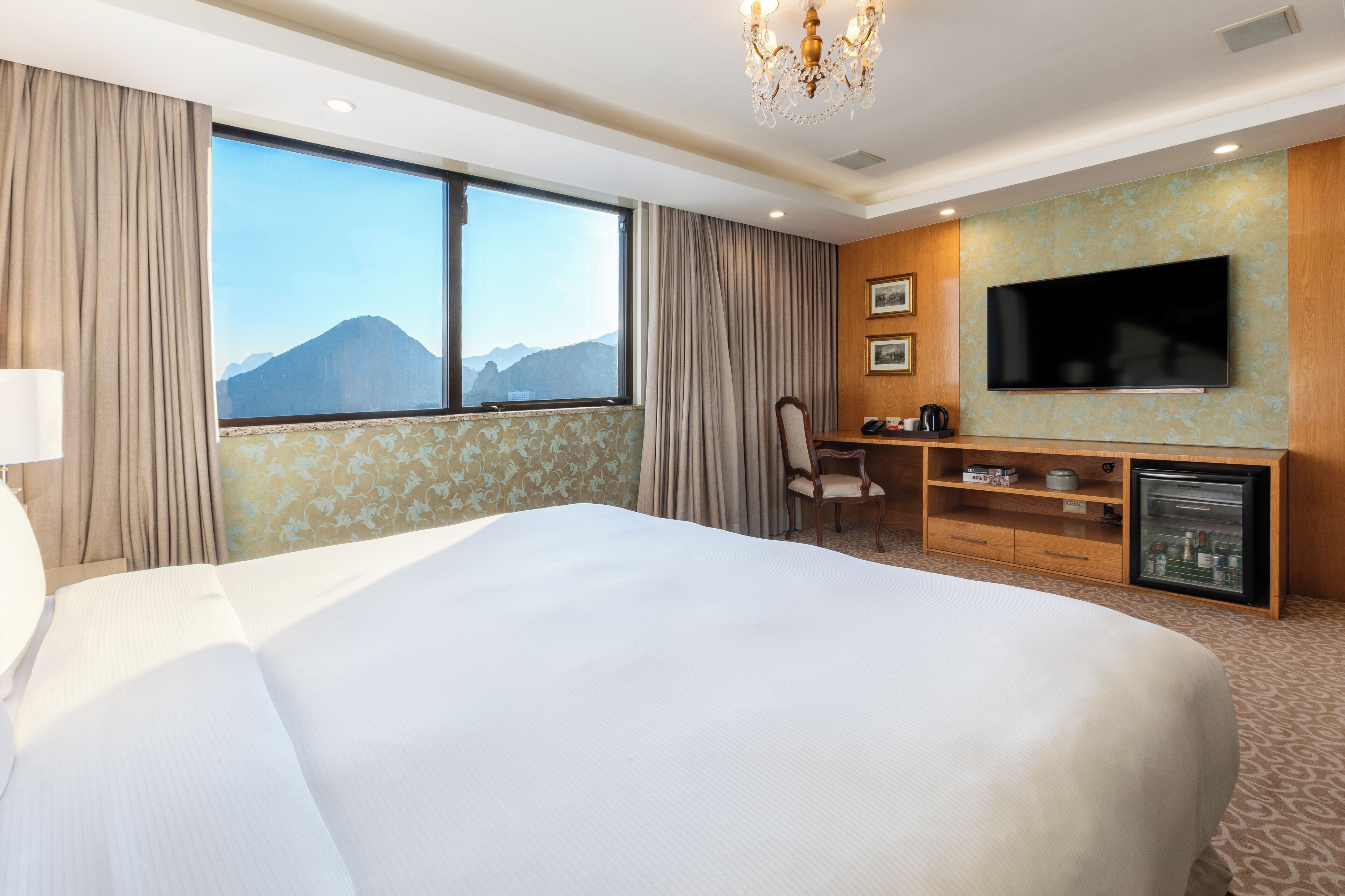 Quarto de hotel com uma cama grande, televisão, escrivaninha com cadeira, frigobar e vista para montanhas pela janela.