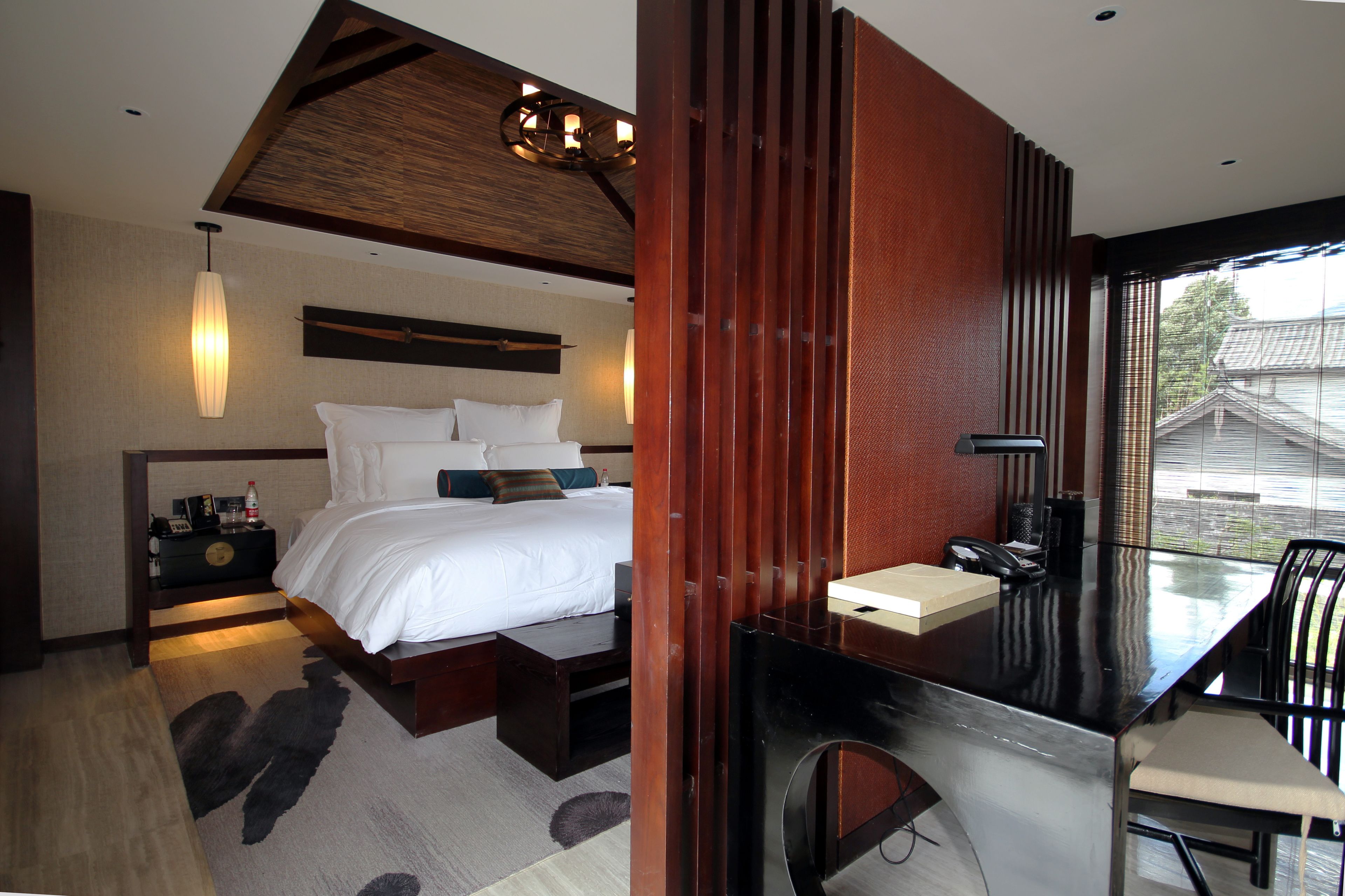 Quarto de hotel com cama, mesa de cabeceira, luminária e escrivaninha com cadeira.