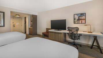Quarto de hotel com duas camas, uma escrivaninha com computador, uma cadeira, uma luminária de mesa e um quadro na parede.