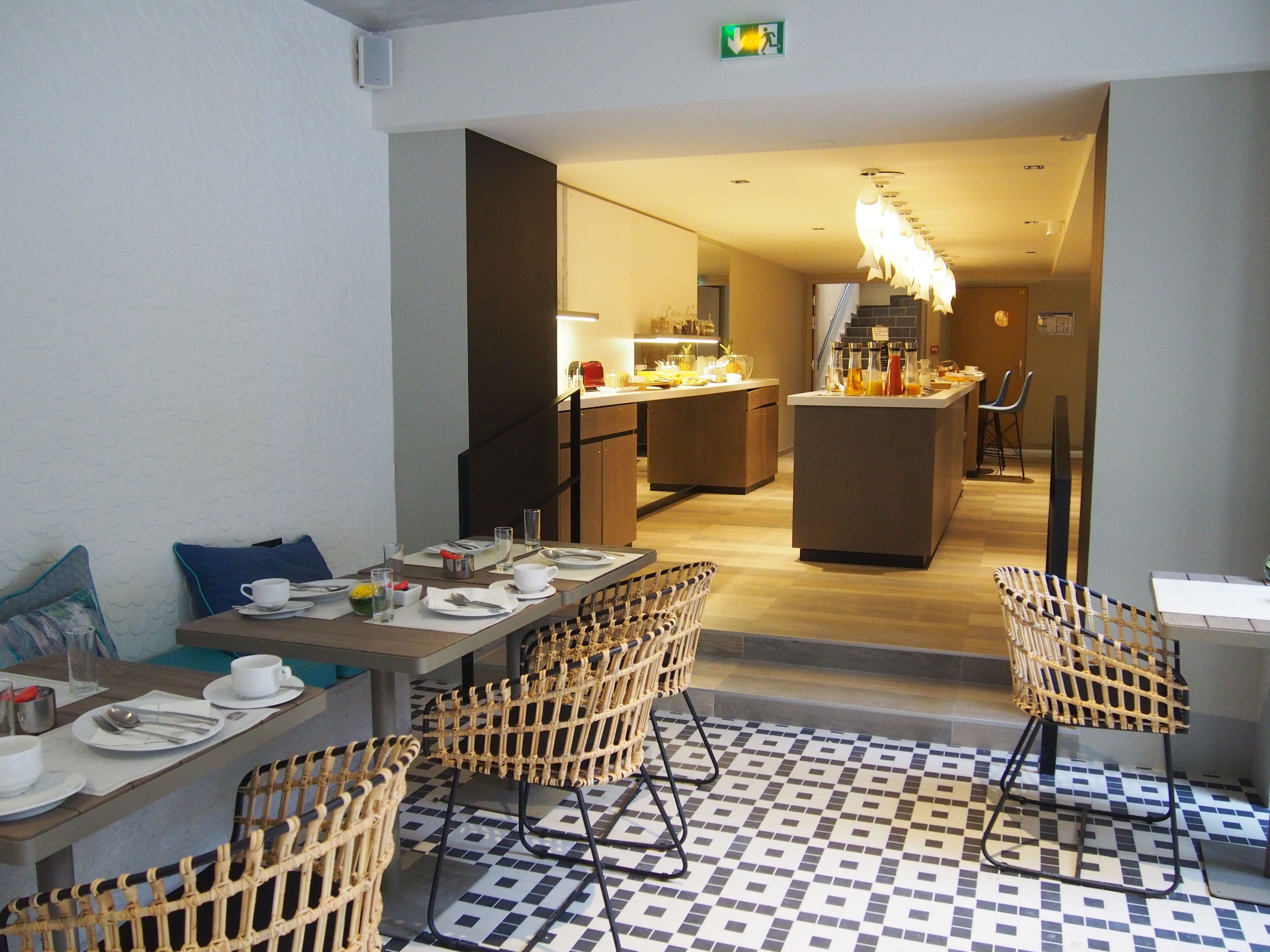 Uma área de refeições de hotel com mesas postas para café da manhã, cadeiras de vime e piso ladrilhado.