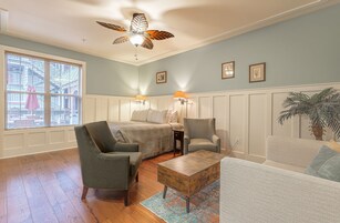 Quarto de hotel com cama, duas poltronas, mesa de centro de madeira, janela grande com persianas, ventilador de teto e luminárias fixadas na parede.