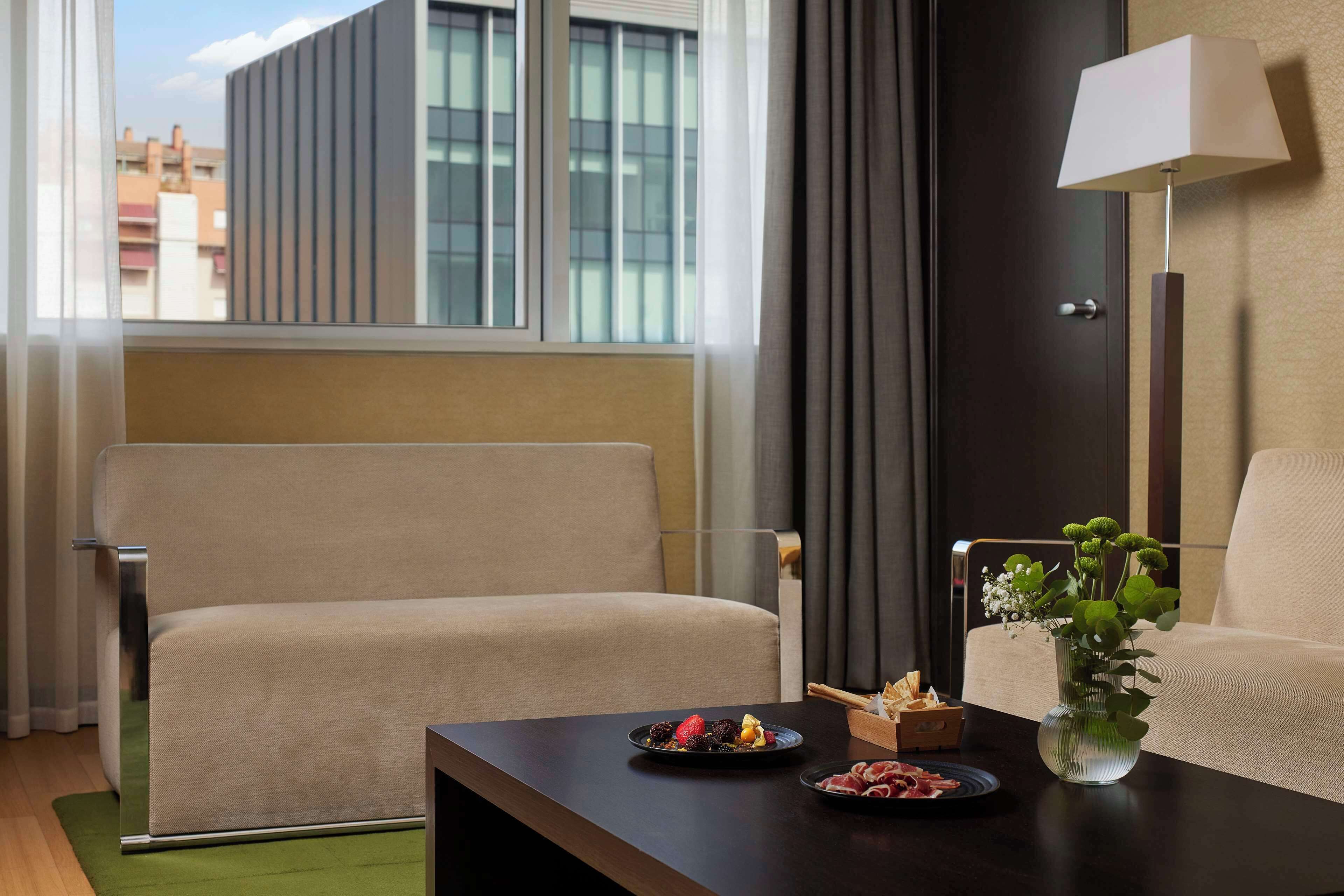 Quarto de hotel moderno com sofá, mesa de jantar com prato de comida, vaso com flores e luminária.