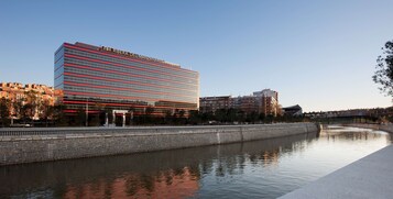 Um edifício moderno com listras horizontais vermelhas e pretas, uma parede de pedra ao lado de um corpo d'água calmo, e um céu claro.