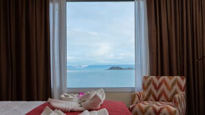Quarto de hotel com cama, poltrona e janela com vista para o mar e montanhas.
