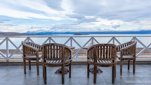 Terraço com cadeiras e mesa de madeira, com vista para uma cordilheira nevada e um corpo d'água.
