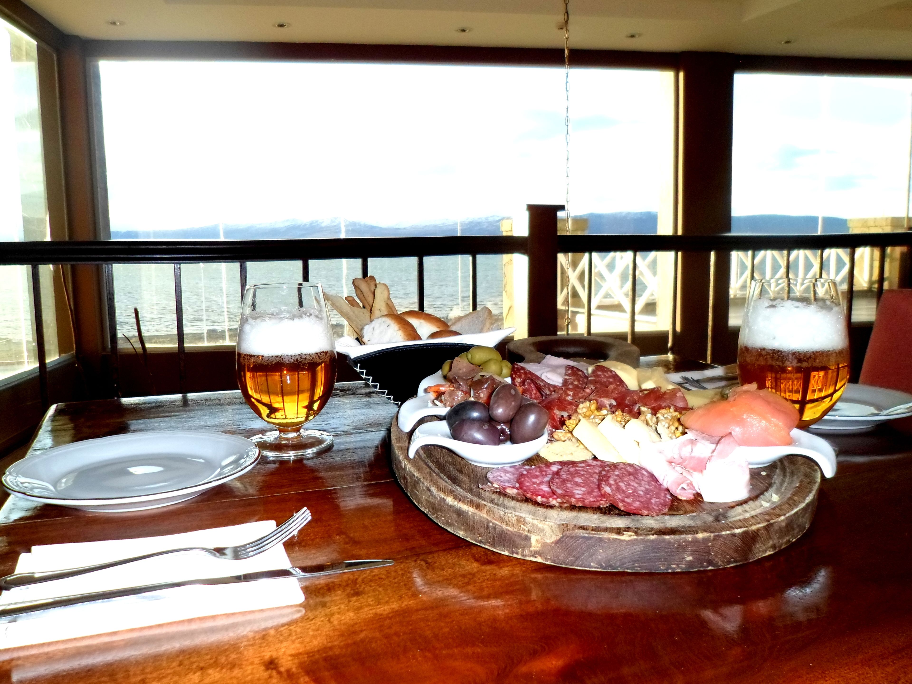 Uma mesa posta com uma tábua de frios, duas canecas de cerveja e vista para o mar.