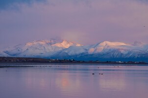 Um lago calmo com montanhas cobertas de neve ao fundo e alguns edifícios na margem.