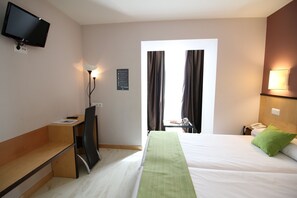 Quarto de hotel com cama, escrivaninha, televisão, abajur, telefone e janela com cortinas.