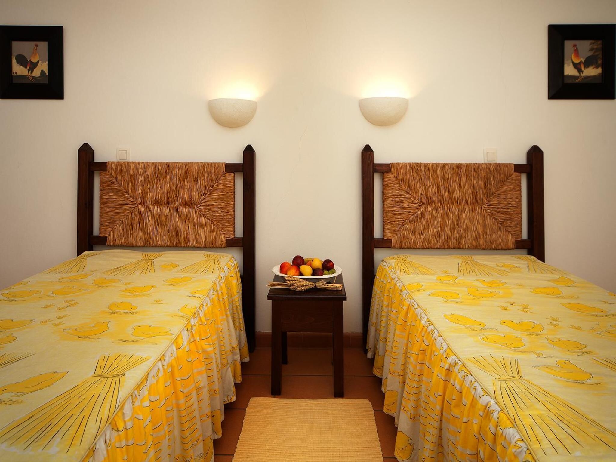 Quarto com duas camas individuais, cada uma com cabeceira de fibra e edredom amarelo com padrão amarelo. Há uma pequena mesa com uma cesta de frutas no centro do quarto.