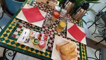 Uma mesa com toalha xadrez, um coador de café, um termo, um copo de suco de laranja, um prato de pão e diversos acompanhamentos.