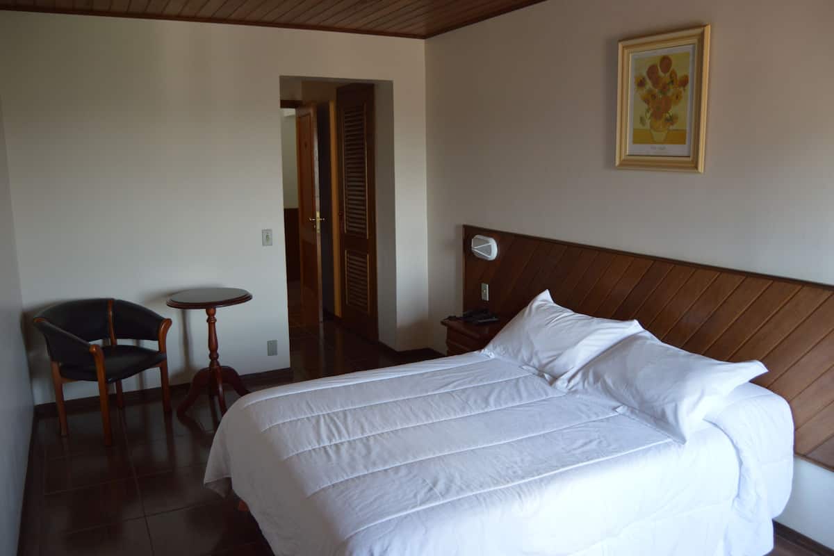 Quarto de hotel com uma cama grande, uma mesa pequena, uma cadeira e uma porta que dá para outro quarto.