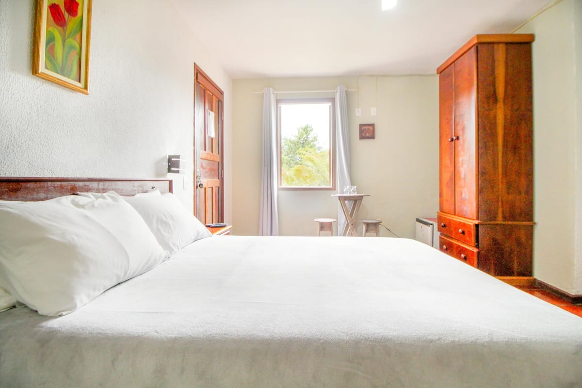 Quarto com uma cama grande, um guarda-roupa de madeira, uma mesinha e uma janela com cortinas.