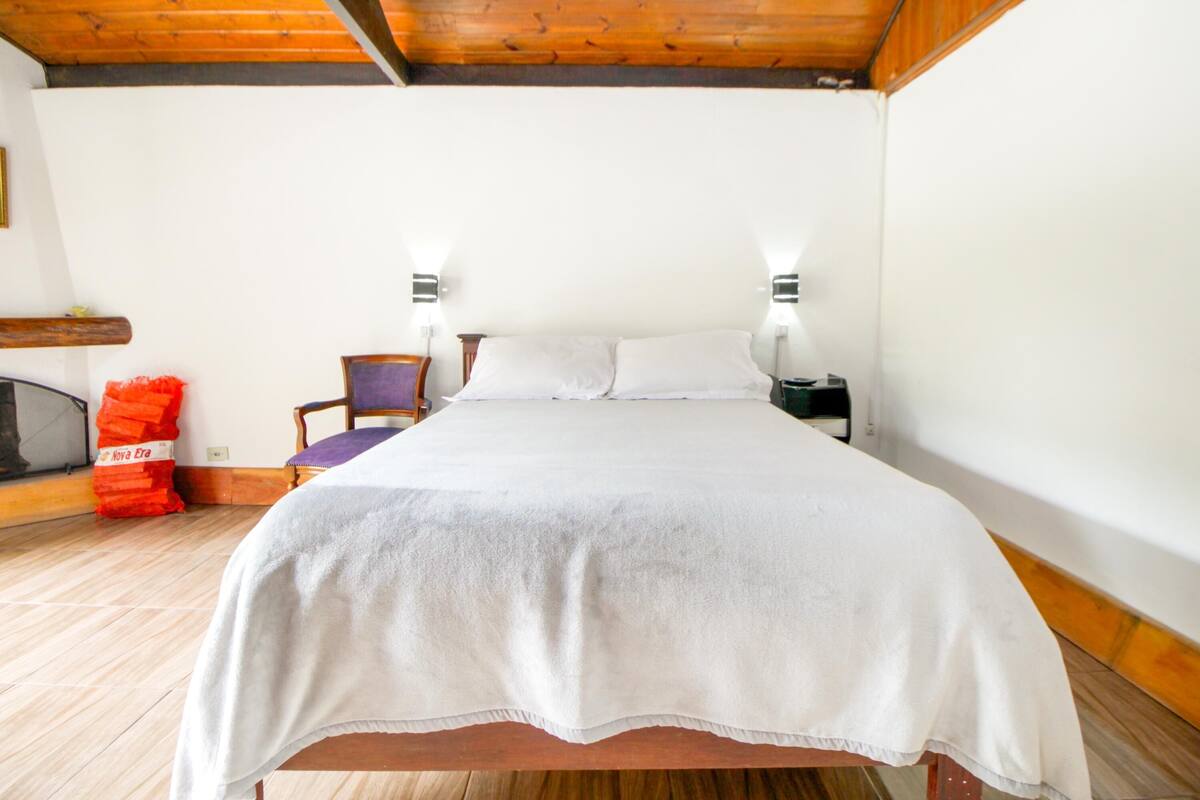 Cama bem arrumada com edredom branco, cabeceira de madeira e duas luminárias de apoio.